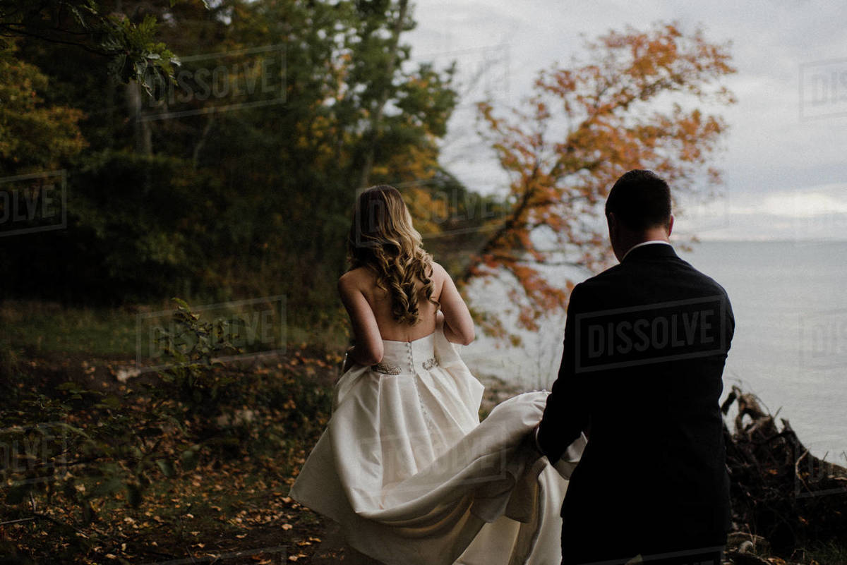 Romantic young groom carrying bride's wedding dress train on lakeside ...