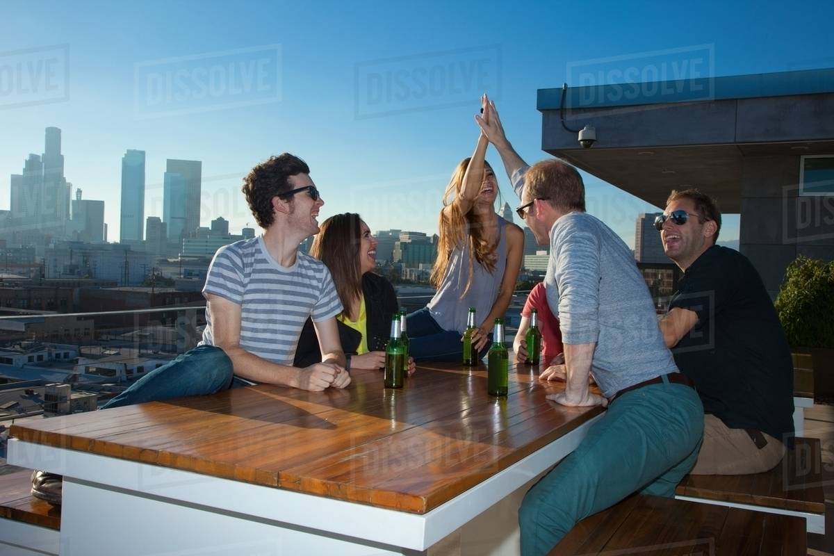 Six adult friends drinking beer at table of rooftop bar with Los ...