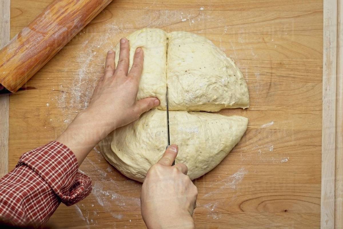 A flatlay photo of a ball of dough being cut up on a large wooden cutting board to be rolled out ...