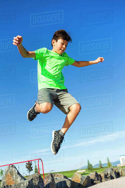 A young boy leaps off a rock in a park on a bright summer day ...