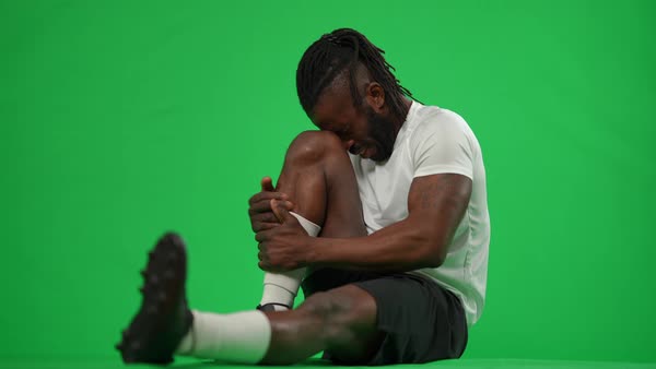 Side view African American footballer sitting on green screen holding ...