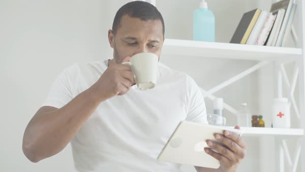A young man sitting in a hospital ward drinking tea and using a tablet ...