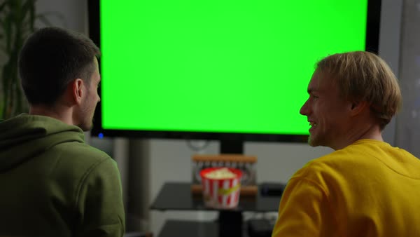 Two positive men talking sitting on couch with chromakey TV screen at ...