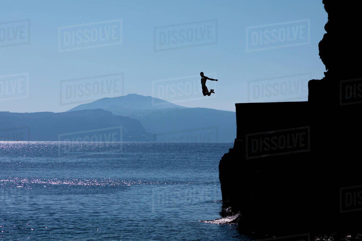 Tourists jumping off a large rock ledge in Amoudi Bay on Santorini ...