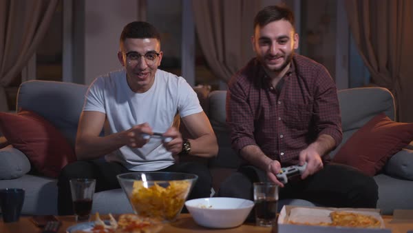 Two friends playing video game and sitting on couch in apartment ...