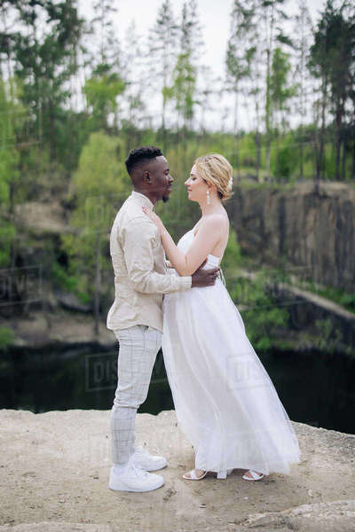 Happy interracial couple newlyweds stands on rock and embraces against ...