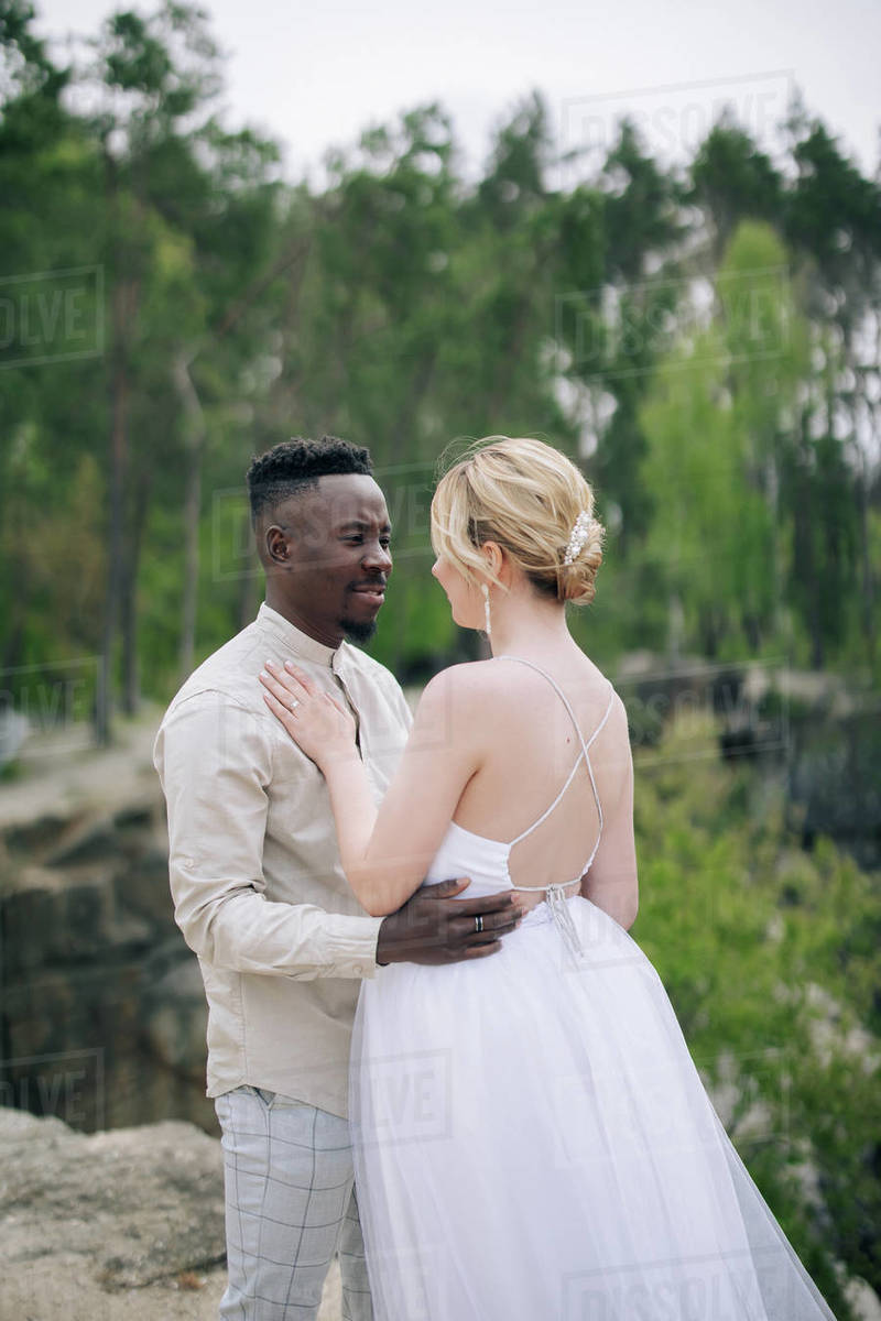Happy interracial couple newlyweds stands on rock and embraces against ...