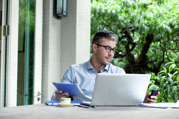 Young white man sitting and working on his laptop holding his cellphone ...