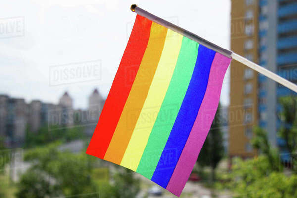 Rainbow LGBT flag set against blue sky, green trees, city street and ...