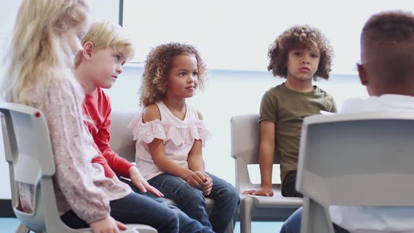 Infant school kids sitting on chairs talking with their teacher in the ...