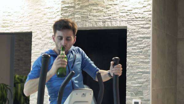 Drunk man exercising on elliptical machine in the gym and drinking beer ...
