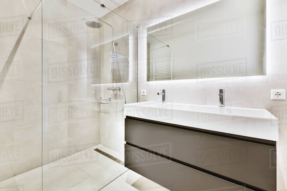Sink with taps and illuminated mirror installed near glass shower wall ...