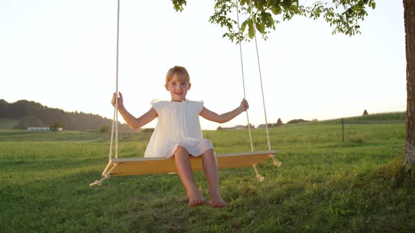 SLOW MOTION CLOSE UP: Smiling little girl swaying on swing at golden ...