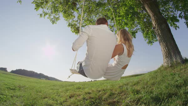 SLOW MOTION: Cheerful young couple swinging on swing under green tree ...