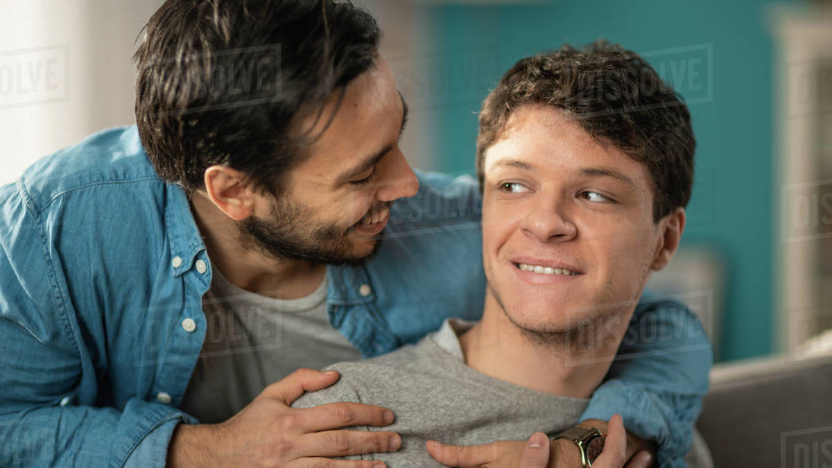 Portrait of a cute male couple at home. Young man sits on a couch, his ...