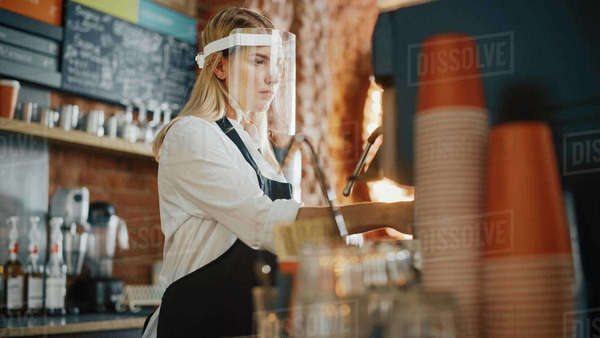 Female barista in a face shield making coffee for a customer while ...