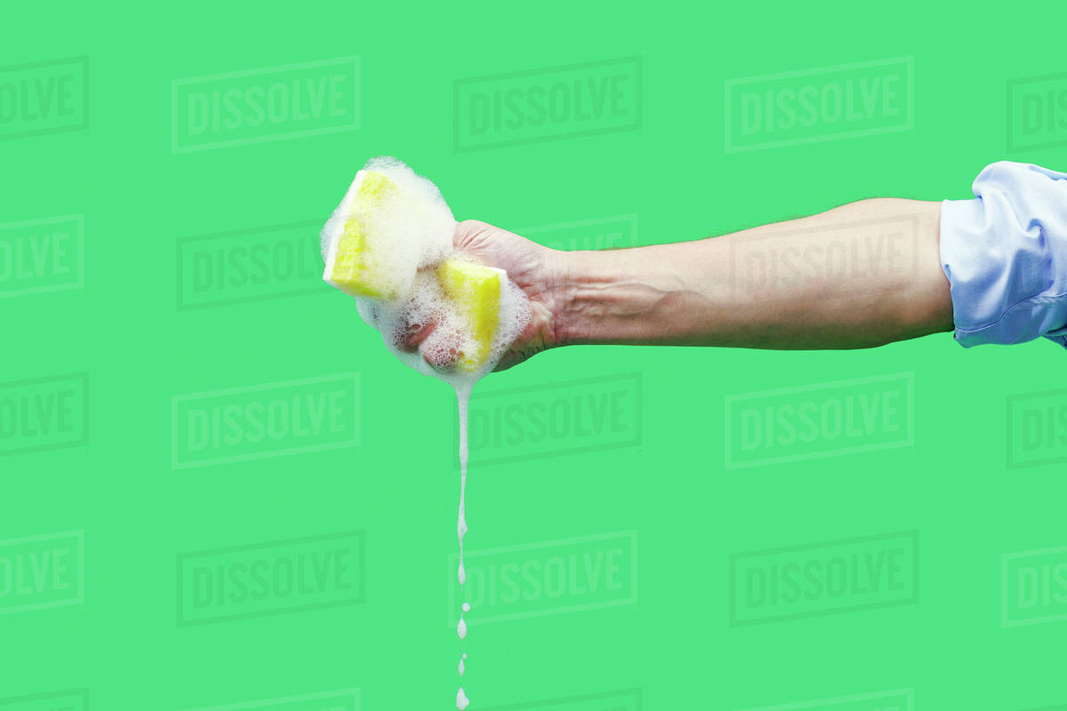Hand squeezing soapy sponge with water dripping on green background ...