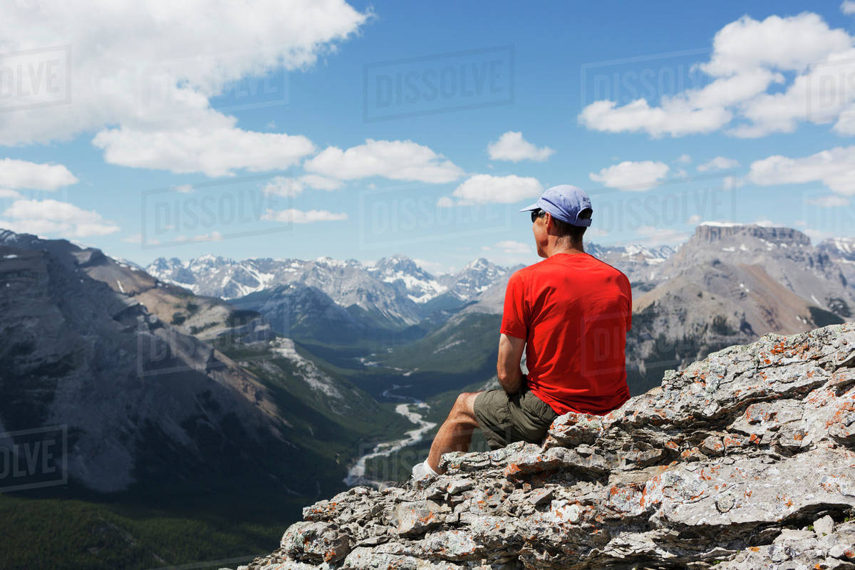Male hiker sitting on a rocky cliff ledge overlooking a river valley ...
