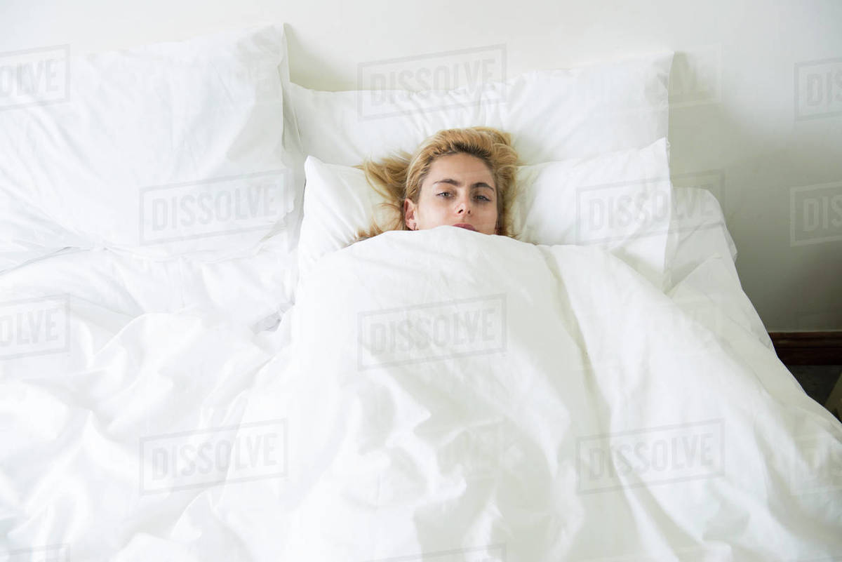 A cool young woman lying in a bed with he covers pulled up to her chin ...