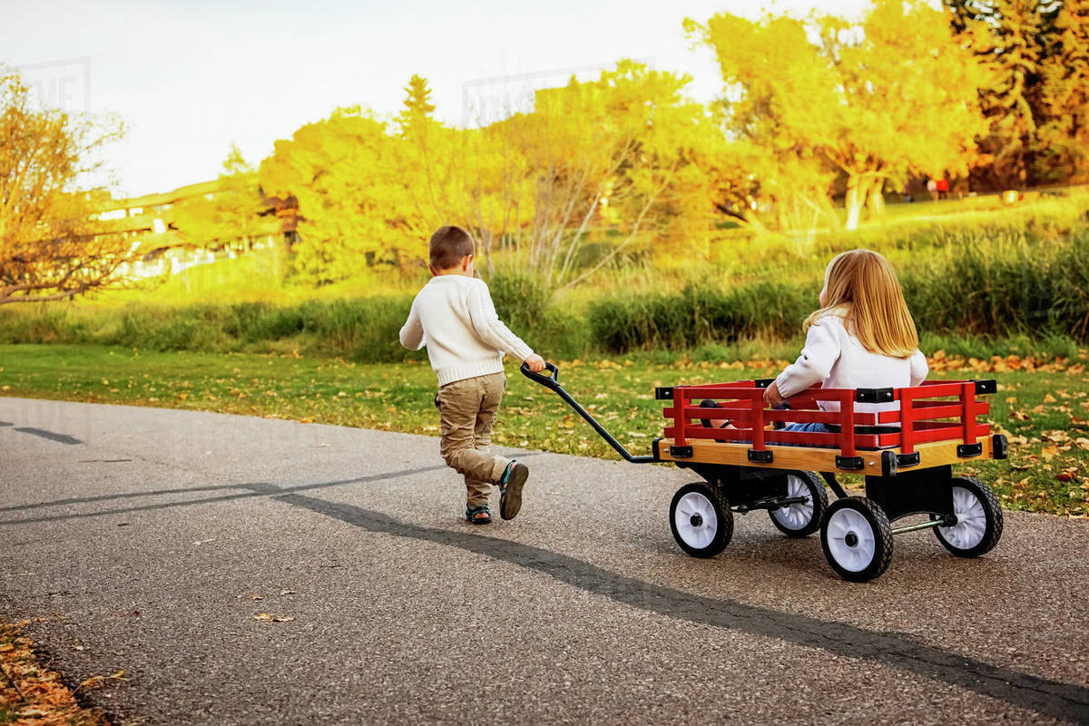 Young boy pulling his sister in a wagon at a city park along a river ...