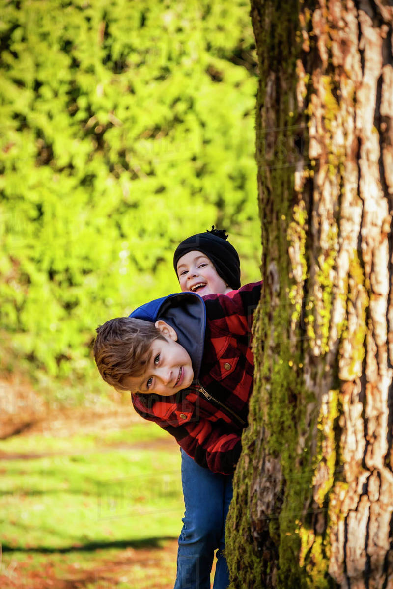 Two young boys peeking out from behind a tree with big smiles on their ...