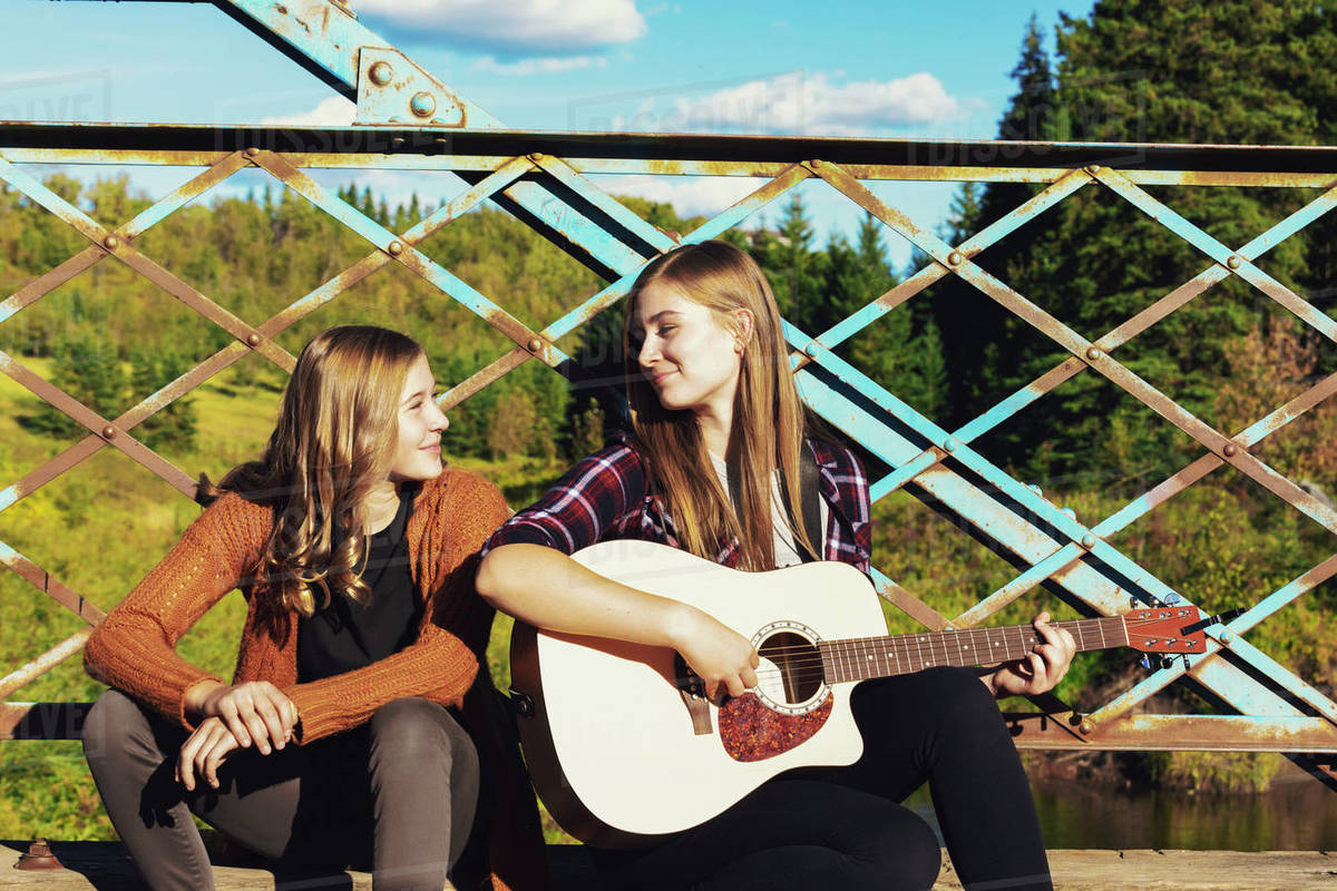 Two sisters sit on a park bridge, one with an acoustic guitar, enjoying ...