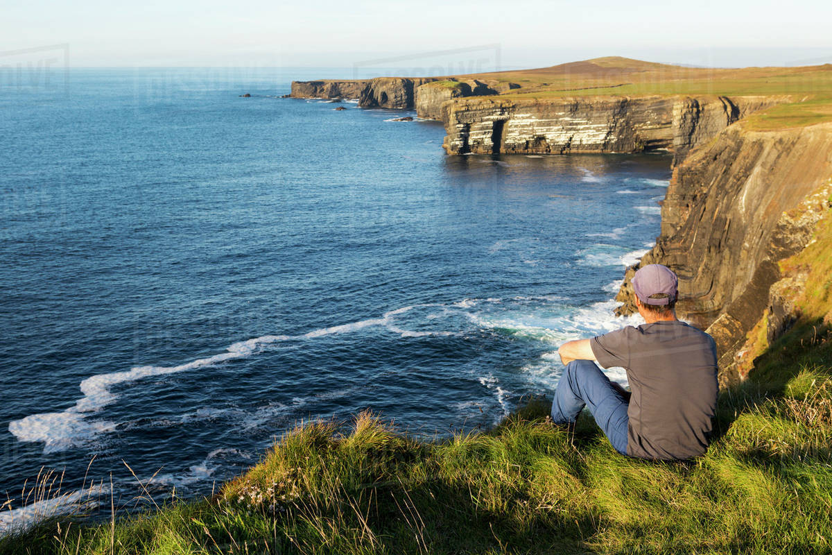 Man sitting on grassy cliff edge overlooking the dramatic rock cliffs ...