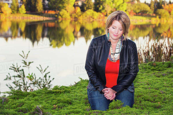 Mature Christian woman kneeling and praying beside a lake in autumn ...