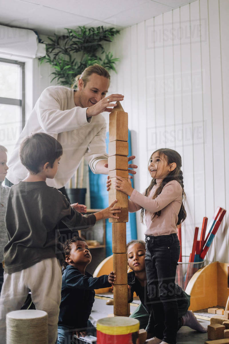 Male teacher guiding children stacking wooden toy blocks at ...