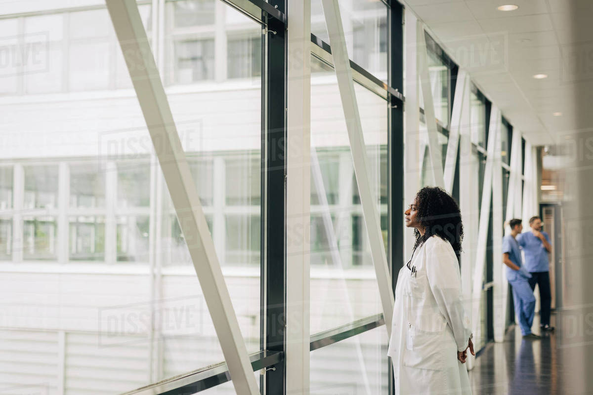 Side view of female physician looking through window while standing in ...