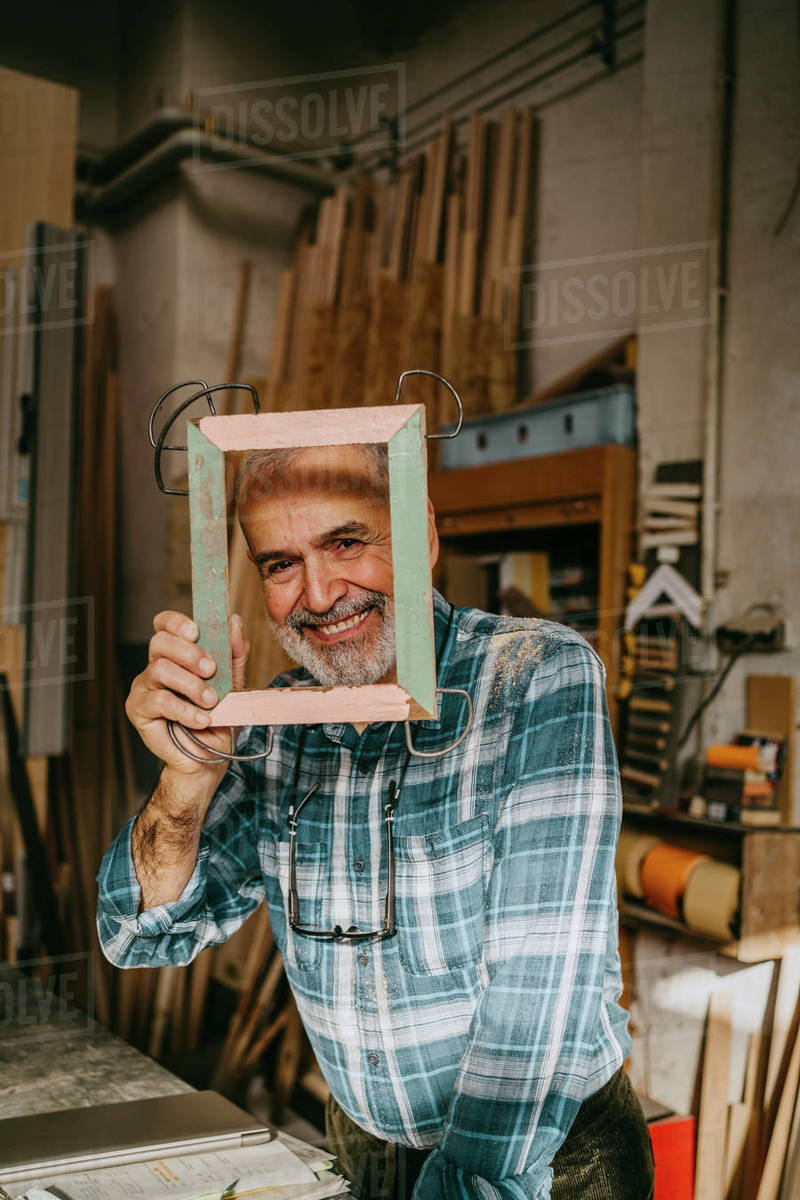 Portrait of happy craftsman looking through picture frame at workshop ...