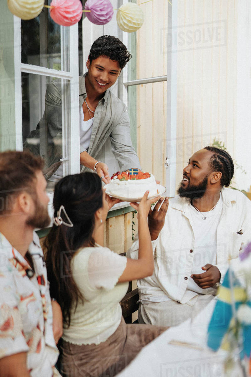 Smiling young man giving cake to male and female friends sitting at ...
