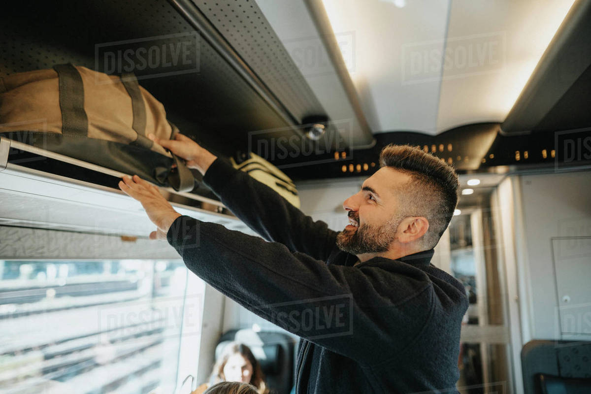 Side view of smiling man loading luggage on shelf in train - Royalty ...