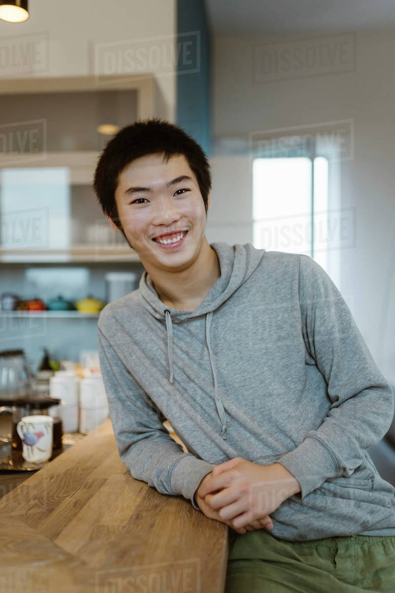 Portrait of smiling teenage boy leaning on wooden counter at home ...