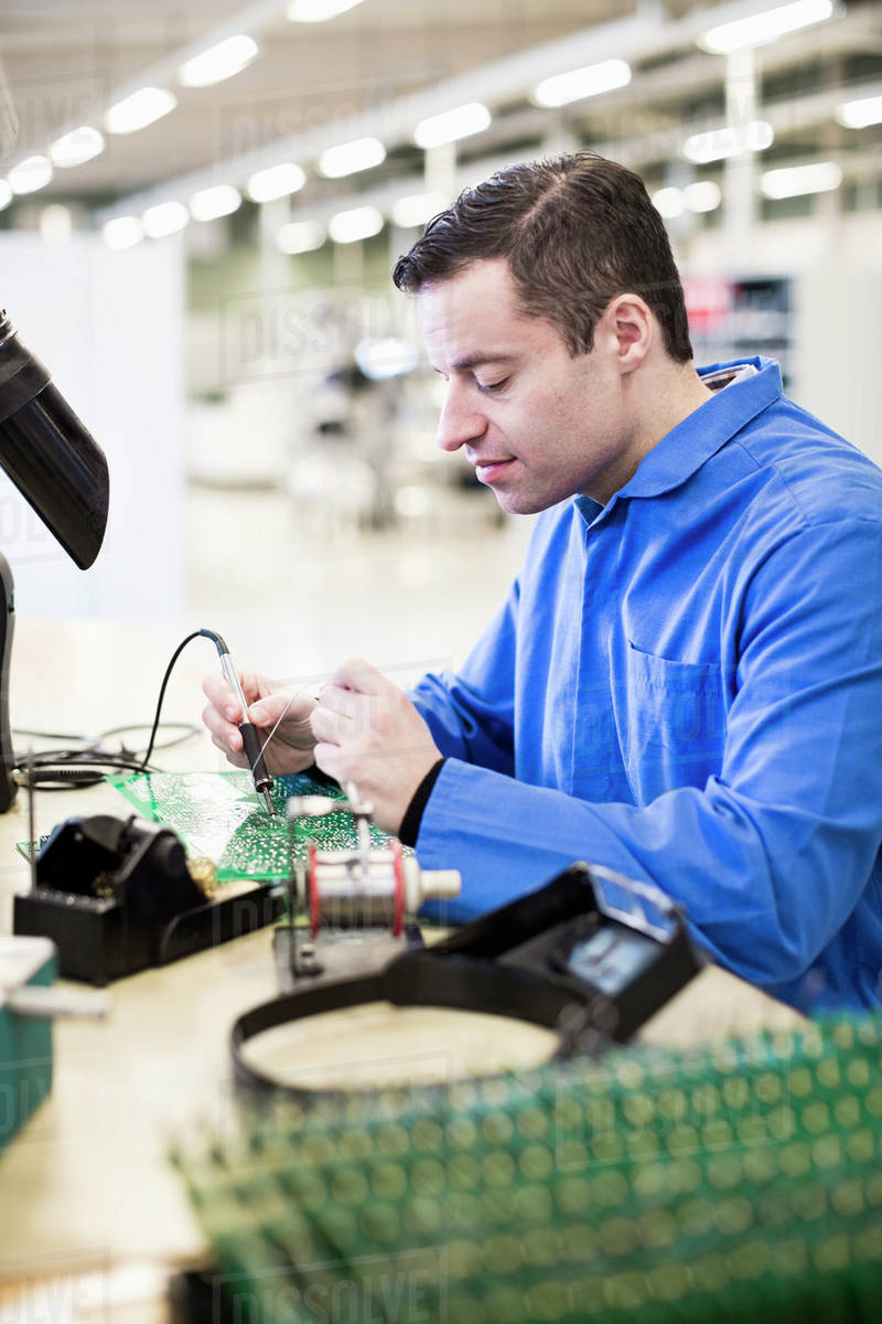 Mature male technician soldering circuit board at desk in industry ...