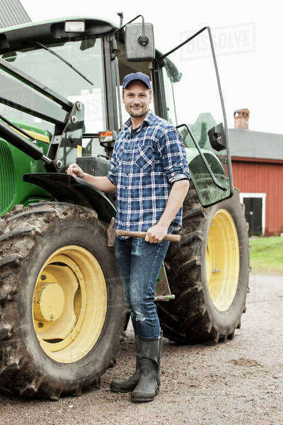 Full length portrait of farmer with hammer standing by tractor on farm ...