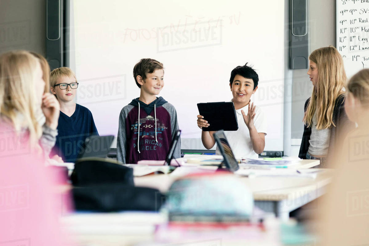 Happy boy giving presentation with digital tablet amidst students in ...