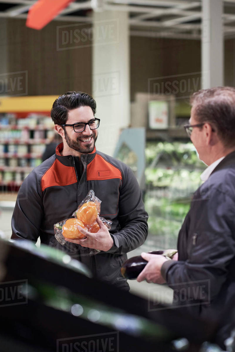 Smiling sales clerk talking man while holding vegetables at supermarket ...
