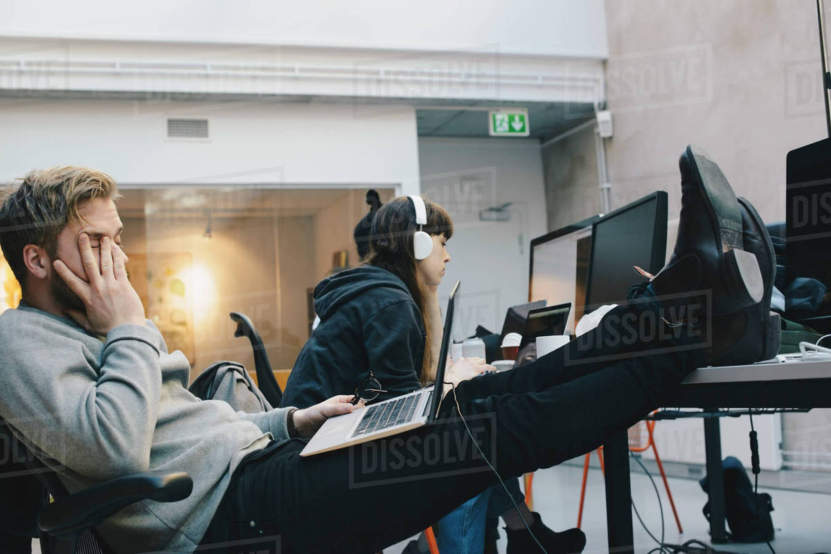 Tired computer programmer sitting with feet up on desk while colleagues ...