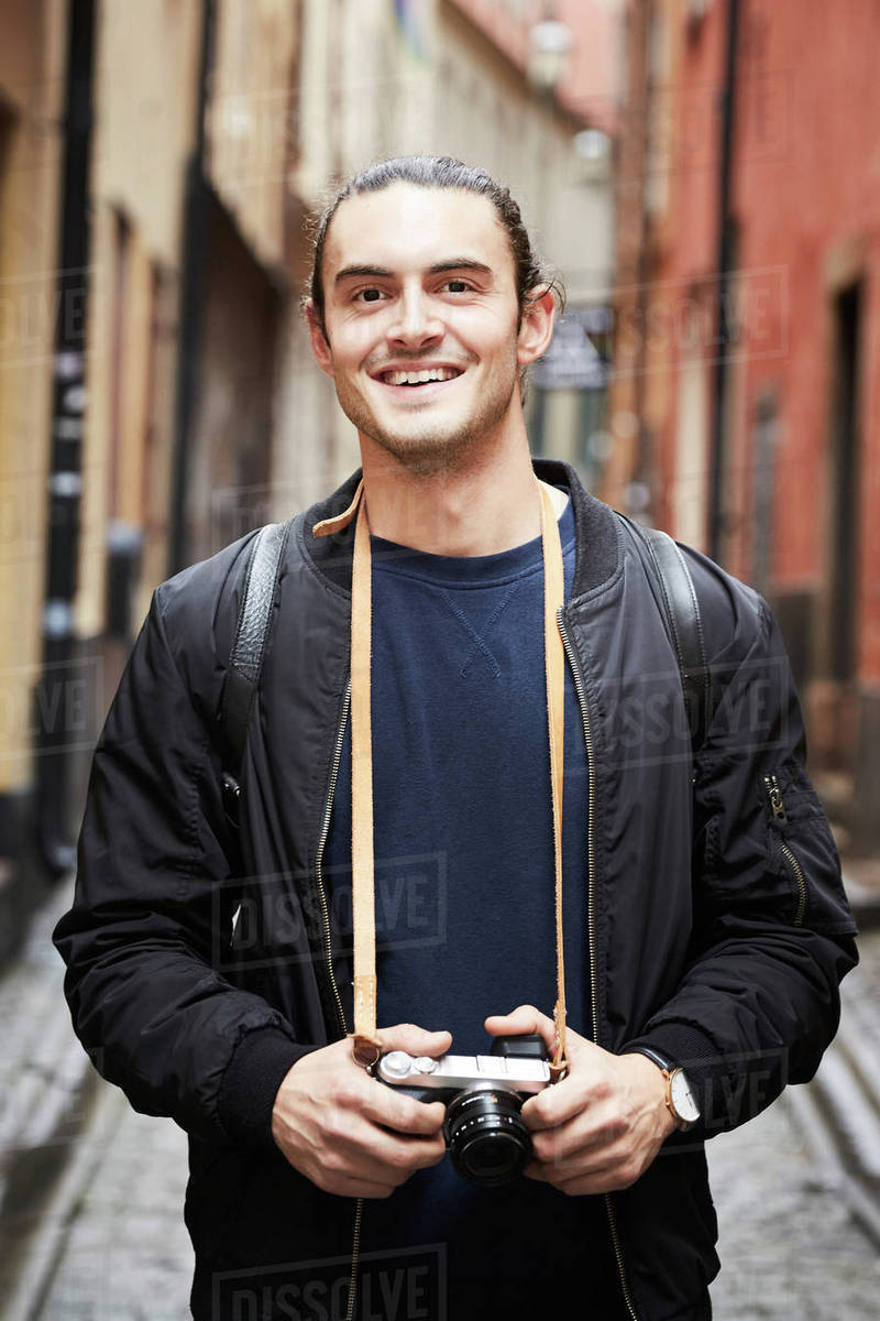 Portrait of smiling young man holding camera while standing in alley ...