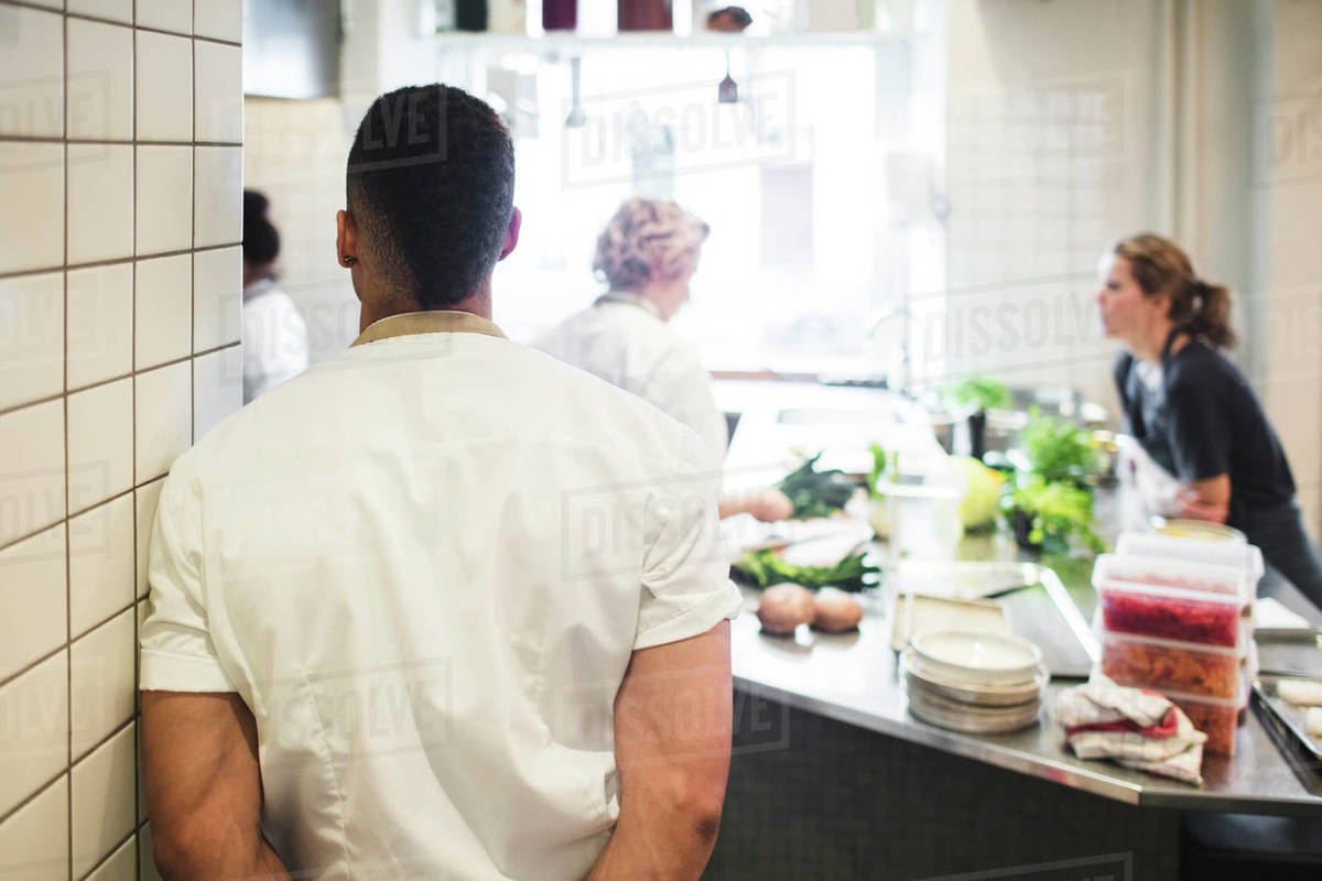 Rear view of male chef standing by wall with female colleagues working ...