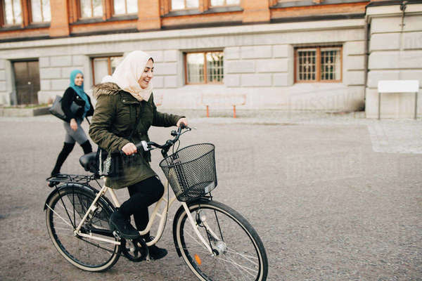 Happy young Muslim woman cycling on street with friend walking in city ...