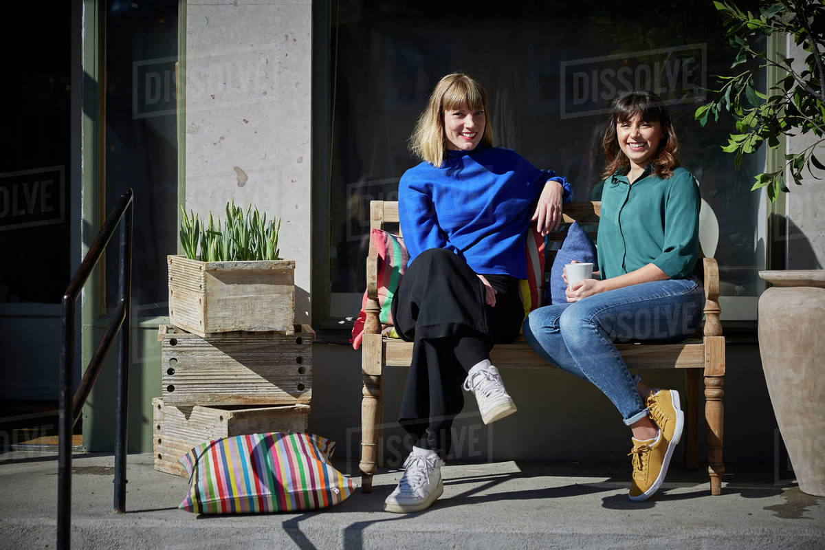 Full length portrait of smiling owners sitting on bench outside store ...