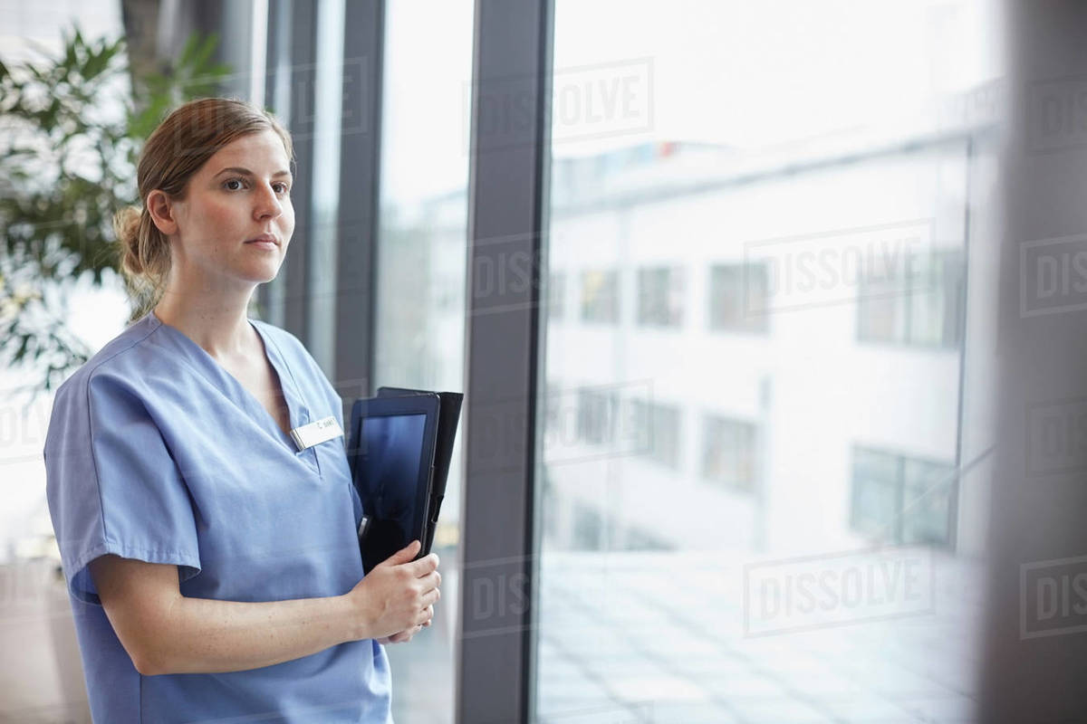 Thoughtful female nurse looking through window while standing in ...