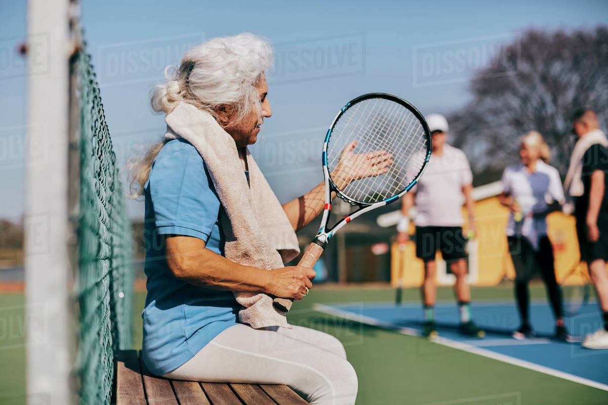 Senior woman holding tennis racket while sitting on bench at tennis ...
