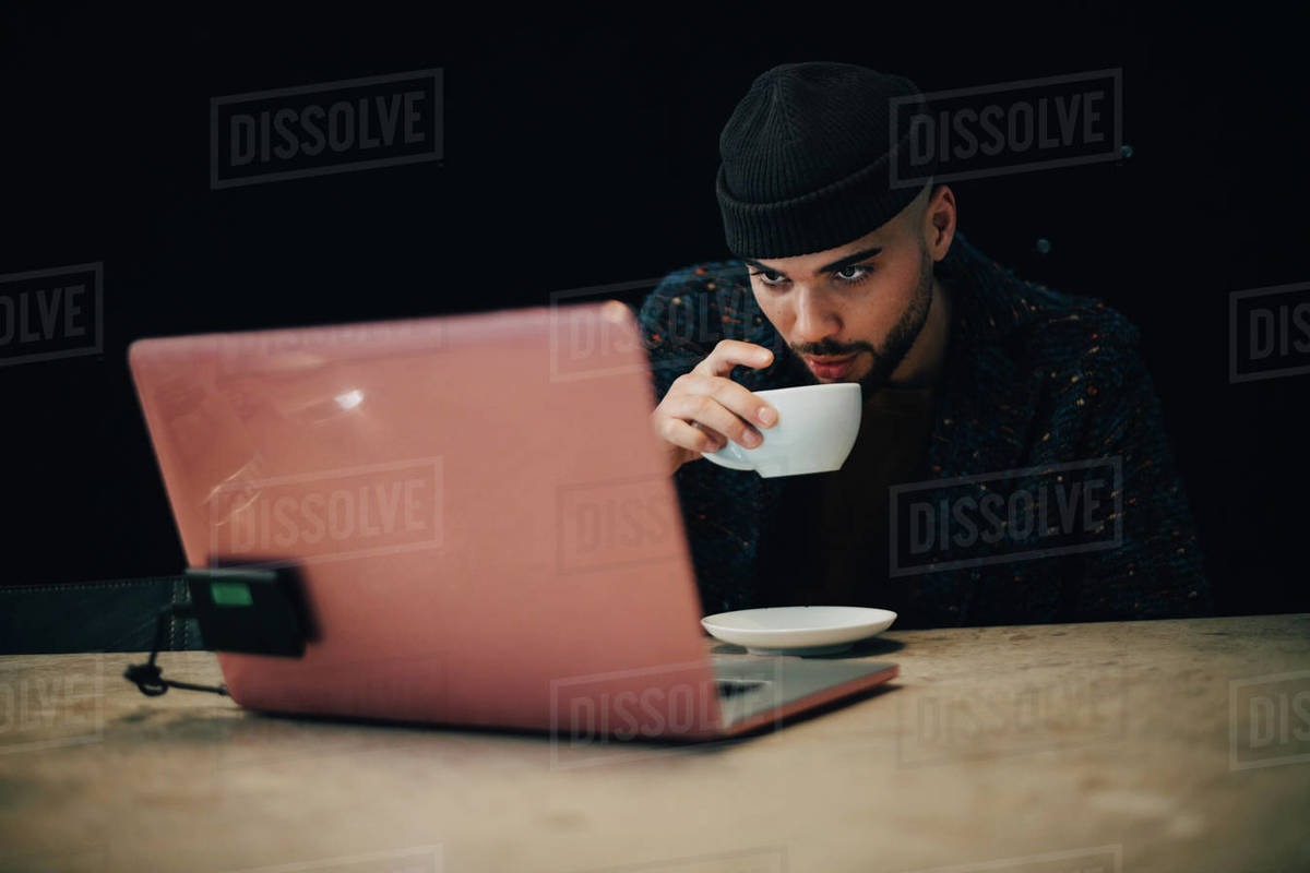 Confident young male hacker drinking coffee while looking at laptop on ...