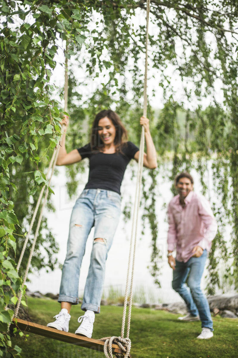 Cheerful woman swinging on rope swing while male friend standing at ...
