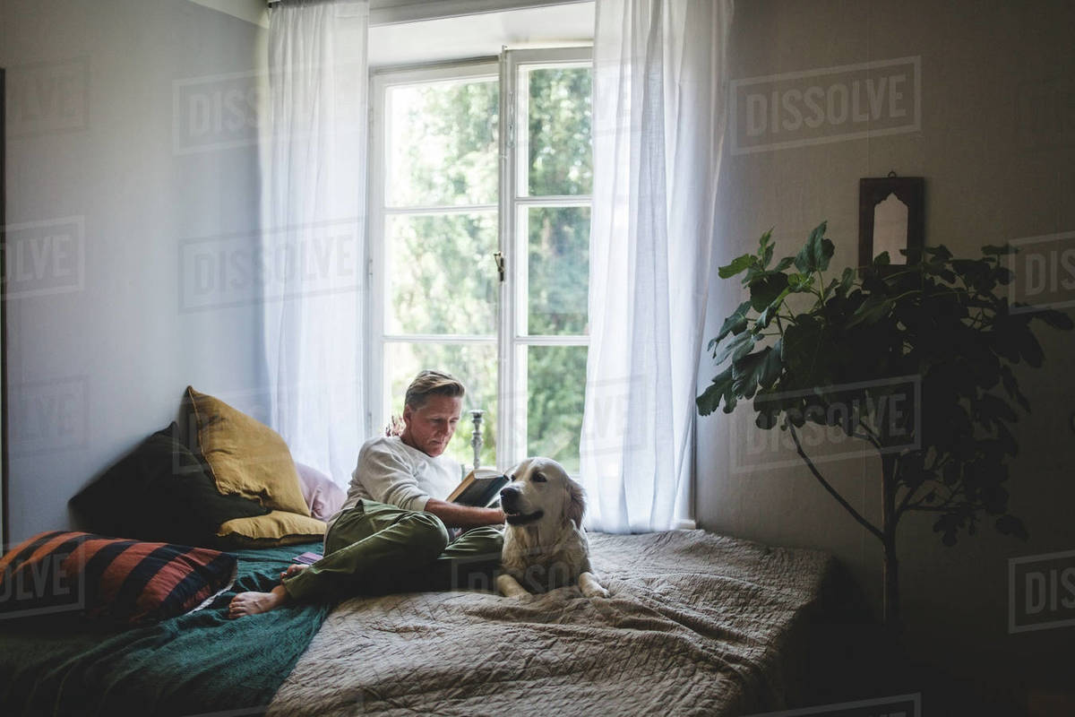 Full length of retired man reading book while resting with dog on bed ...