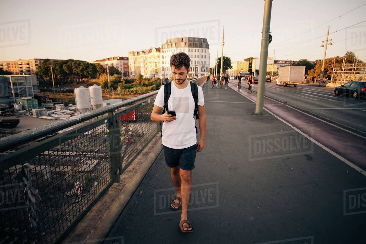 Full length of young man using mobile phone while walking on footpath ...