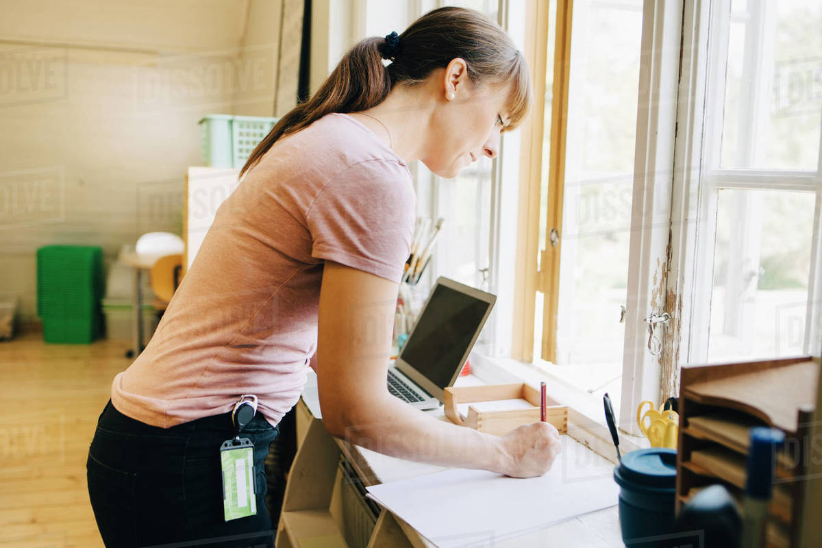 Confident teacher writing on paper while standing by window at ...